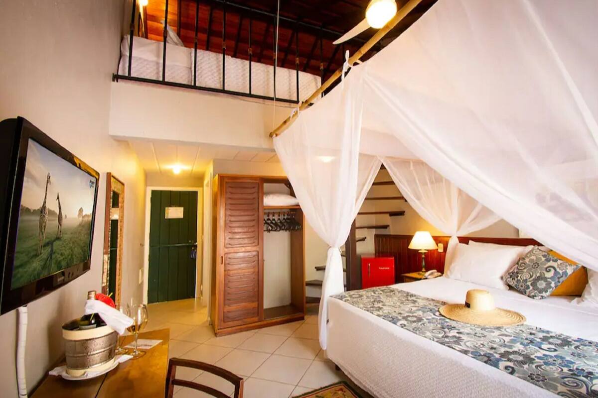Quarto com cama, televisão, escrivaninha com cadeira, guarda-roupa e varanda com mosquiteiro.