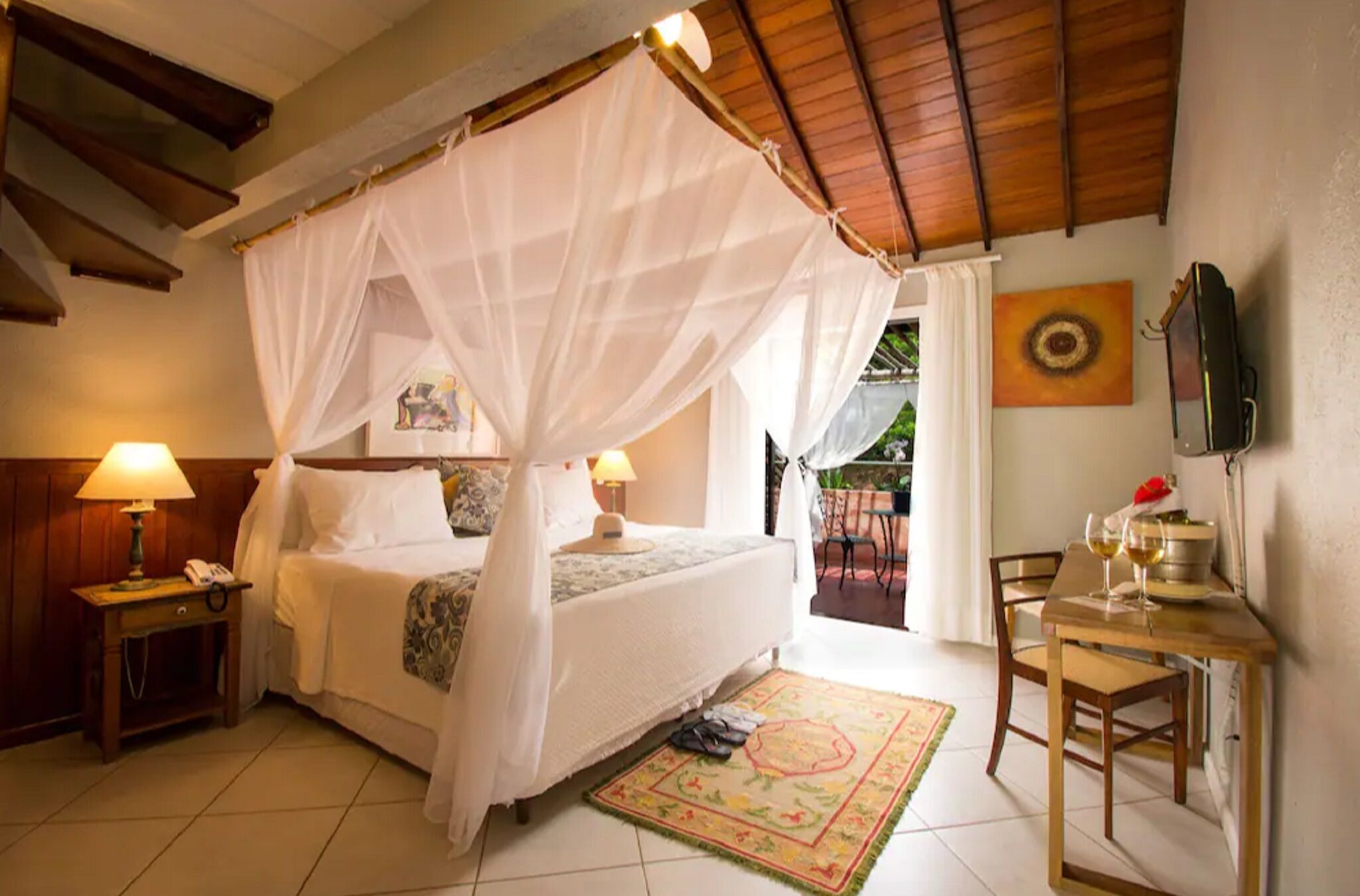 Quarto com cama de dossel, luminárias de cabeceira, uma pequena mesa com cadeira e vista para o exterior.