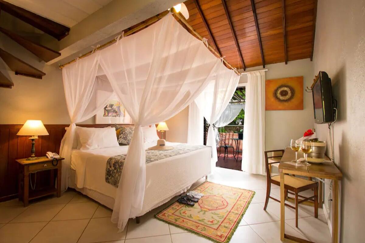 Quarto com cama de dossel, luminárias de cabeceira, uma pequena mesa com cadeira e vista para o exterior.