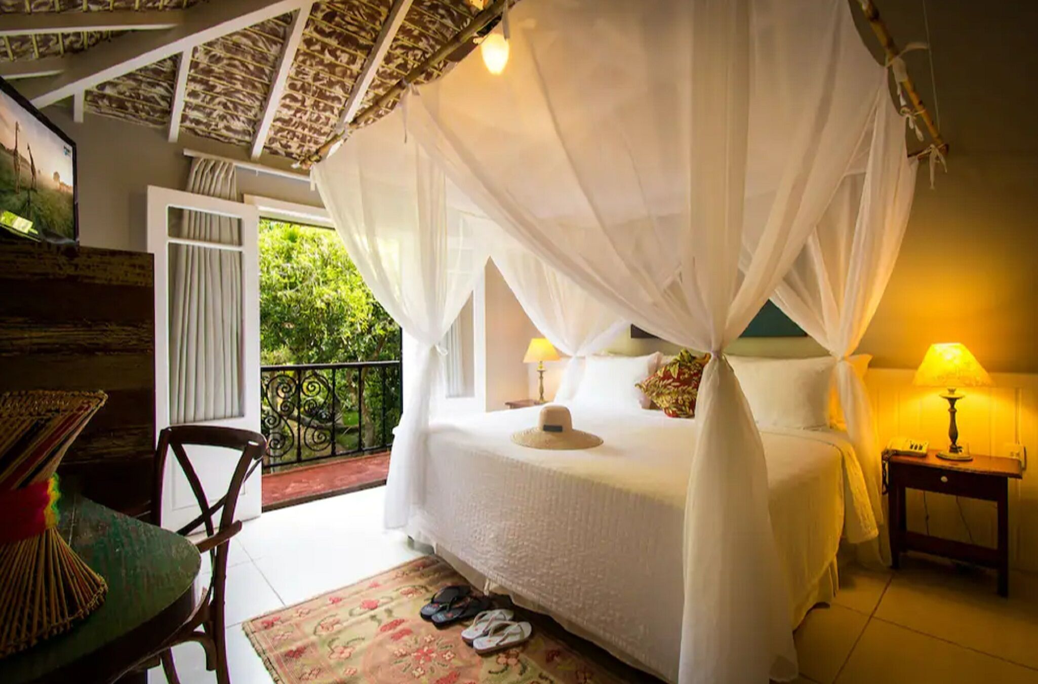 Quarto com cama de dossel, mesa de cabeceira com abajur, uma cadeira e vista para o verde lá fora.