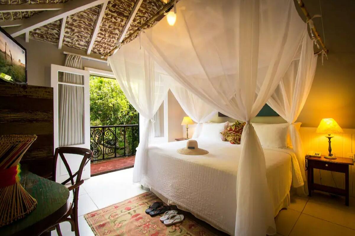 Quarto com cama de dossel, mesa de cabeceira com abajur, uma cadeira e vista para o verde lá fora.