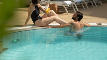 Cena à beira da piscina com uma pessoa sentada na borda e outra na água, um guarda-sol e cadeiras de espreguiçadeira.