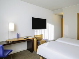 Quarto de hotel com uma cama, uma escrivaninha com cadeira, televisão na parede e janela com persianas.