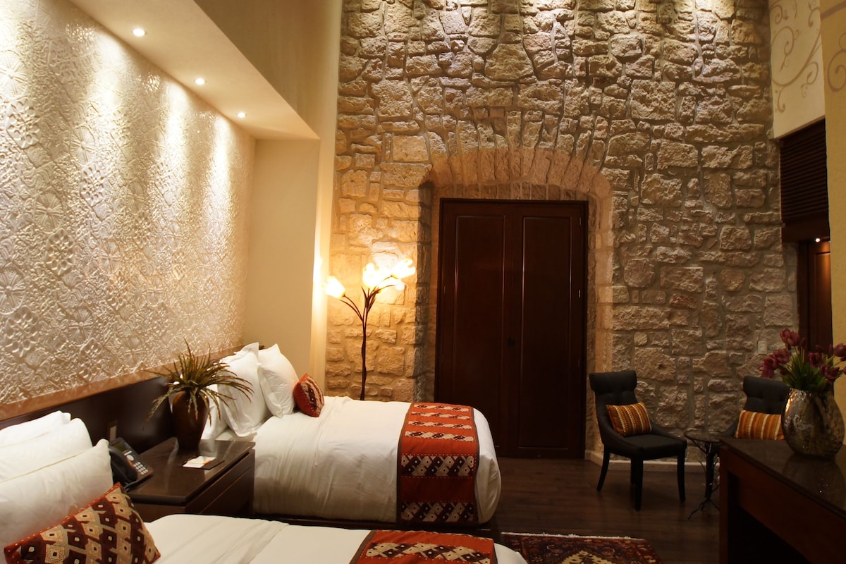 Quarto de hotel com parede de pedra, duas camas com roupa de cama vermelha e branca, teto de madeira e um arco de pedra.