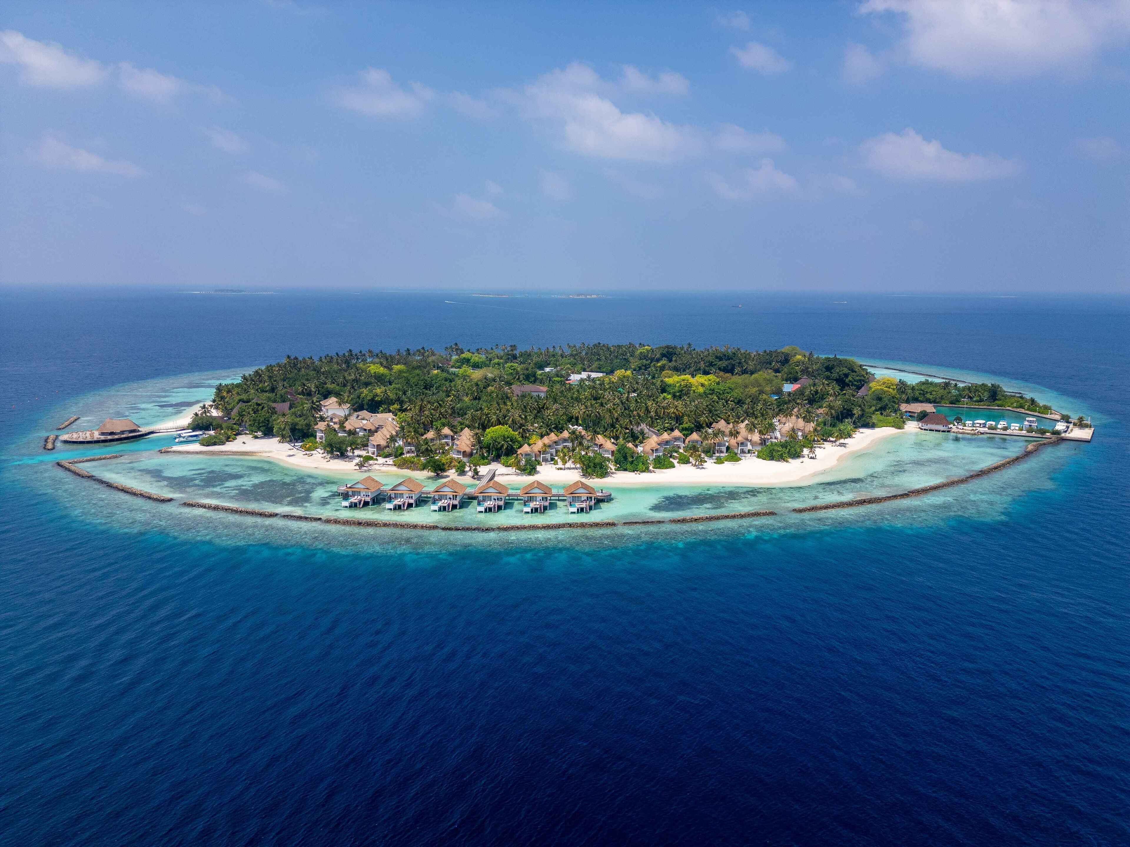 Vista aérea de uma ilha tropical com bangalôs sobre a água, águas turquesa cristalinas e uma área florestada densa.