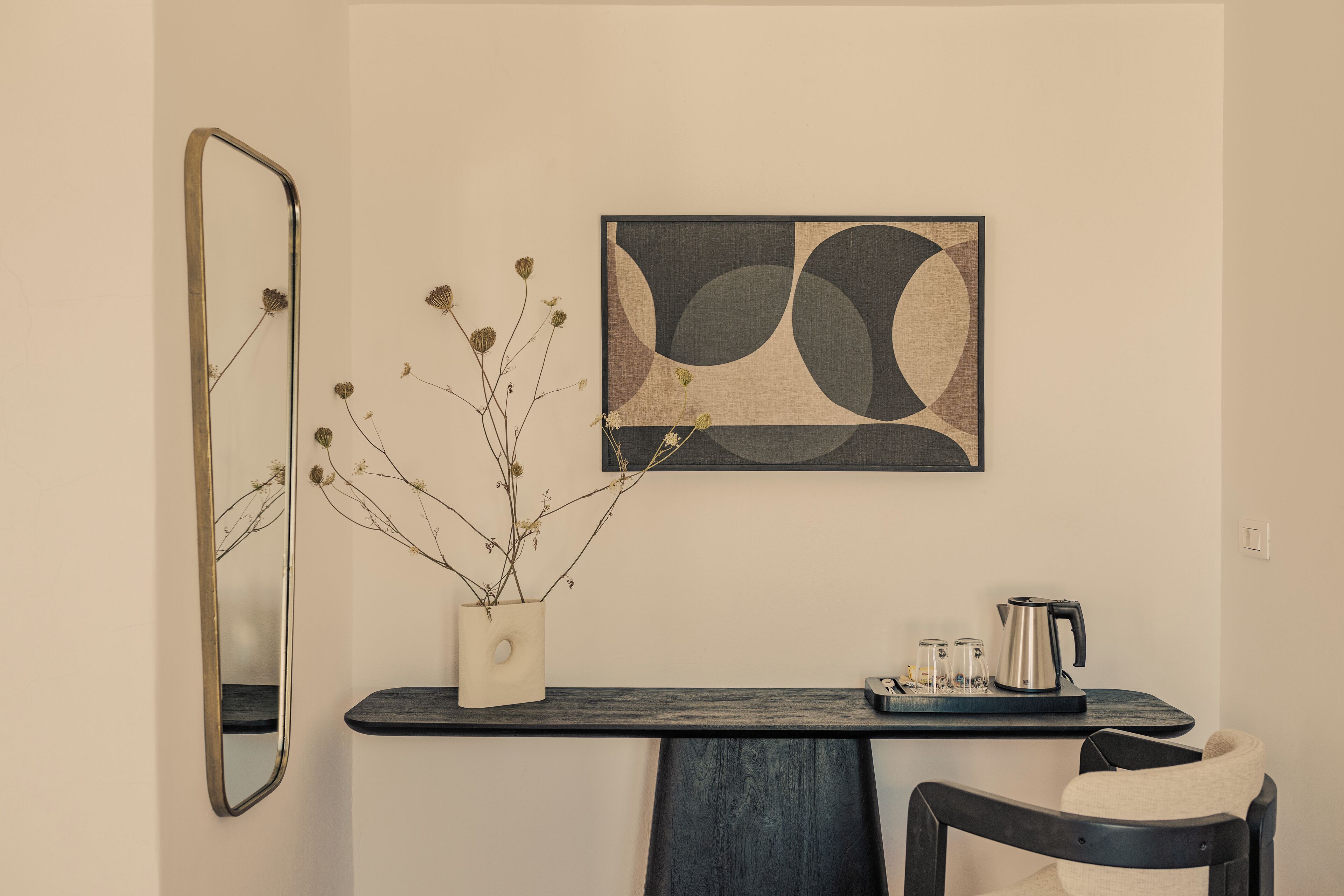 Um banheiro moderno com espelho, um vaso com flores secas, uma pintura abstrata emoldurada e uma pequena mesa com chaleira e copos.