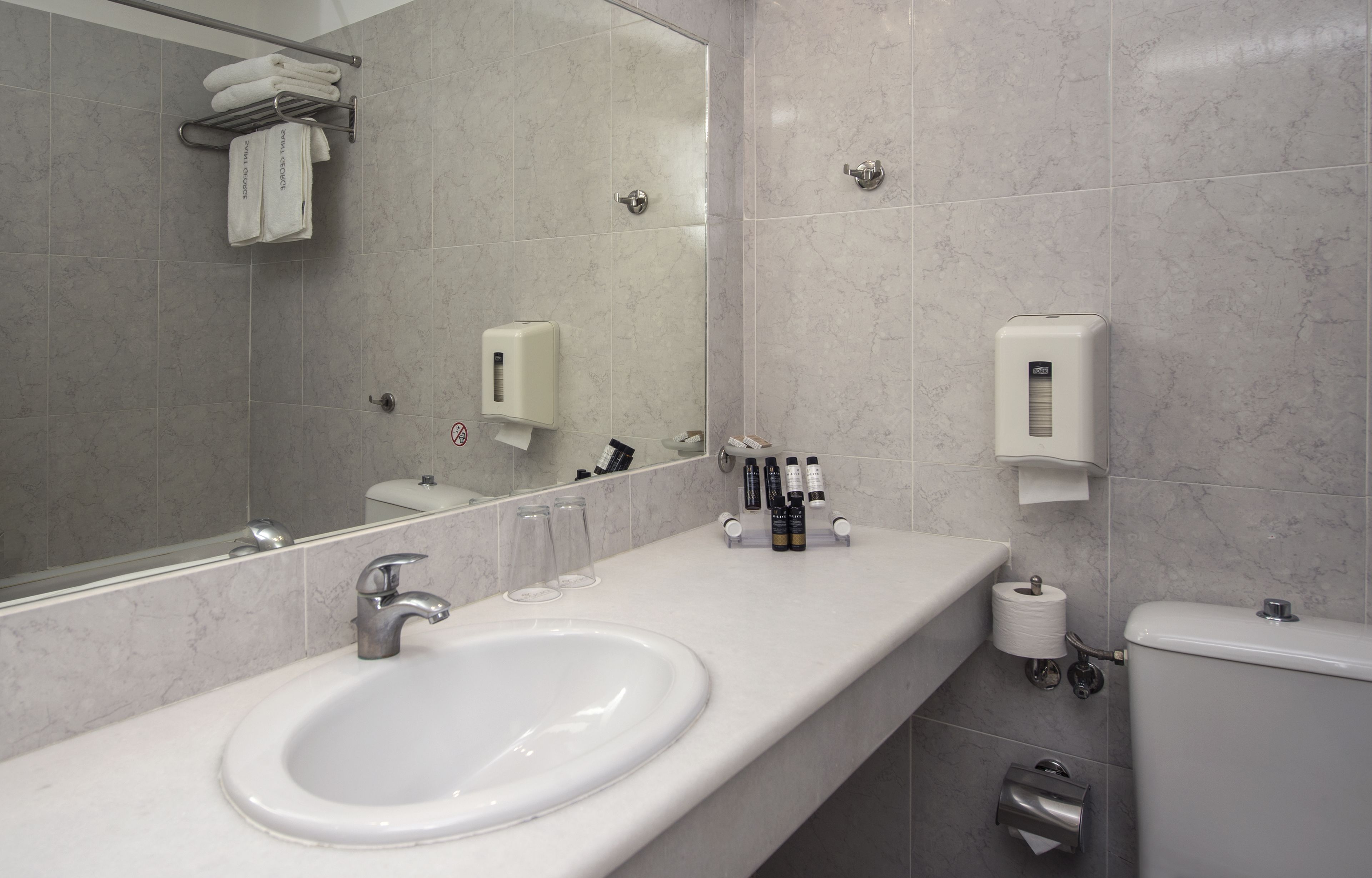 Casa de banho moderna com lavatório branco, espelho e sanita. Há produtos de higiene no balcão, um secador de mãos e um dispensador de papel.
