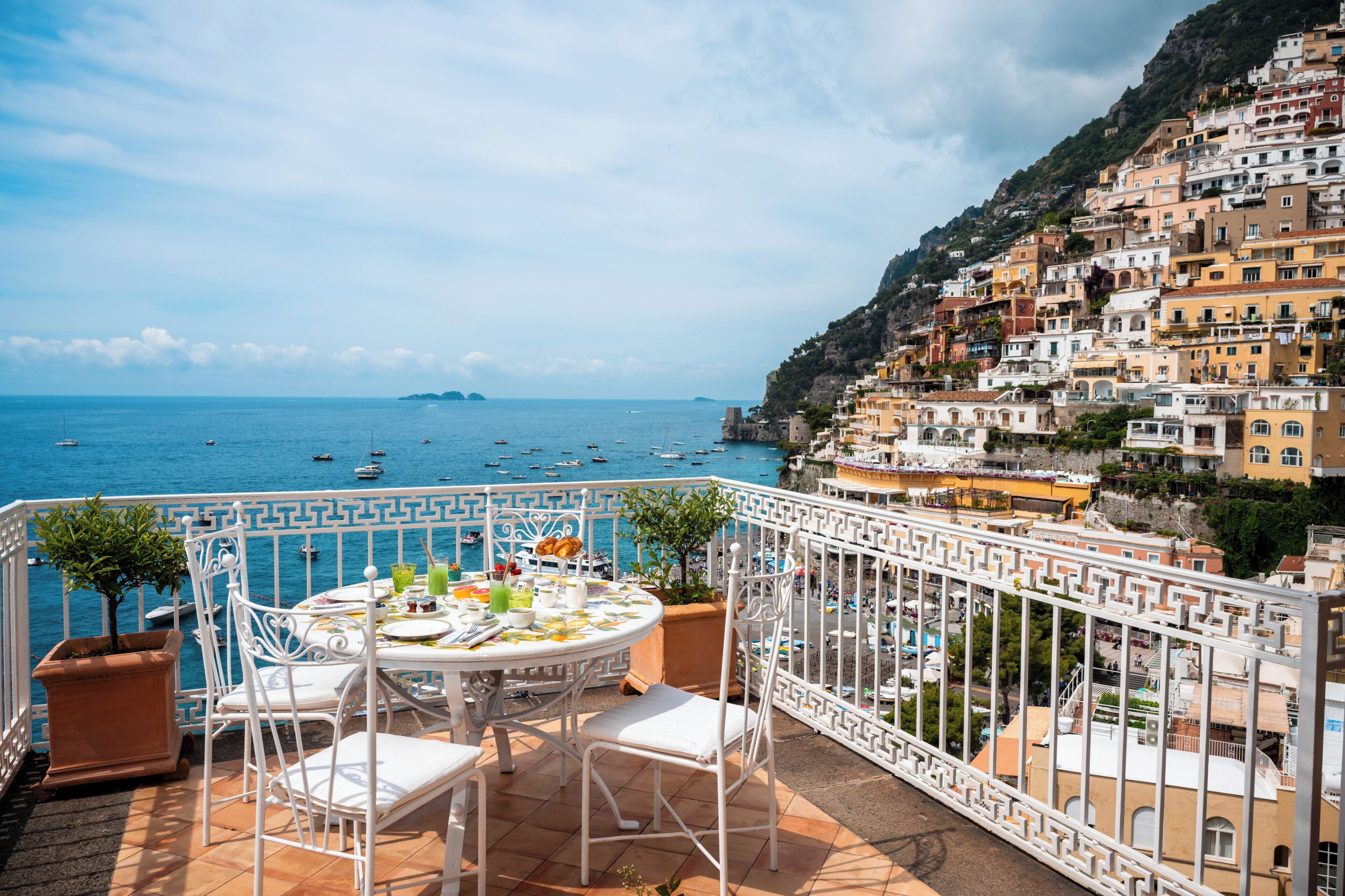 Terraço com mesa posta para refeições, com vista para uma vila costeira e barcos no mar.