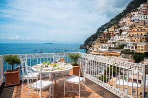 Terraço com mesa posta para refeições, com vista para uma vila costeira e barcos no mar.