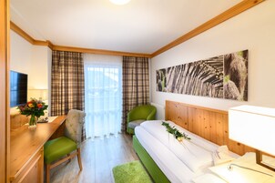 Quarto de hotel com uma escrivaninha de madeira, uma cadeira verde, uma cama com cabeceira branca e uma janela grande com cortinas xadrez.