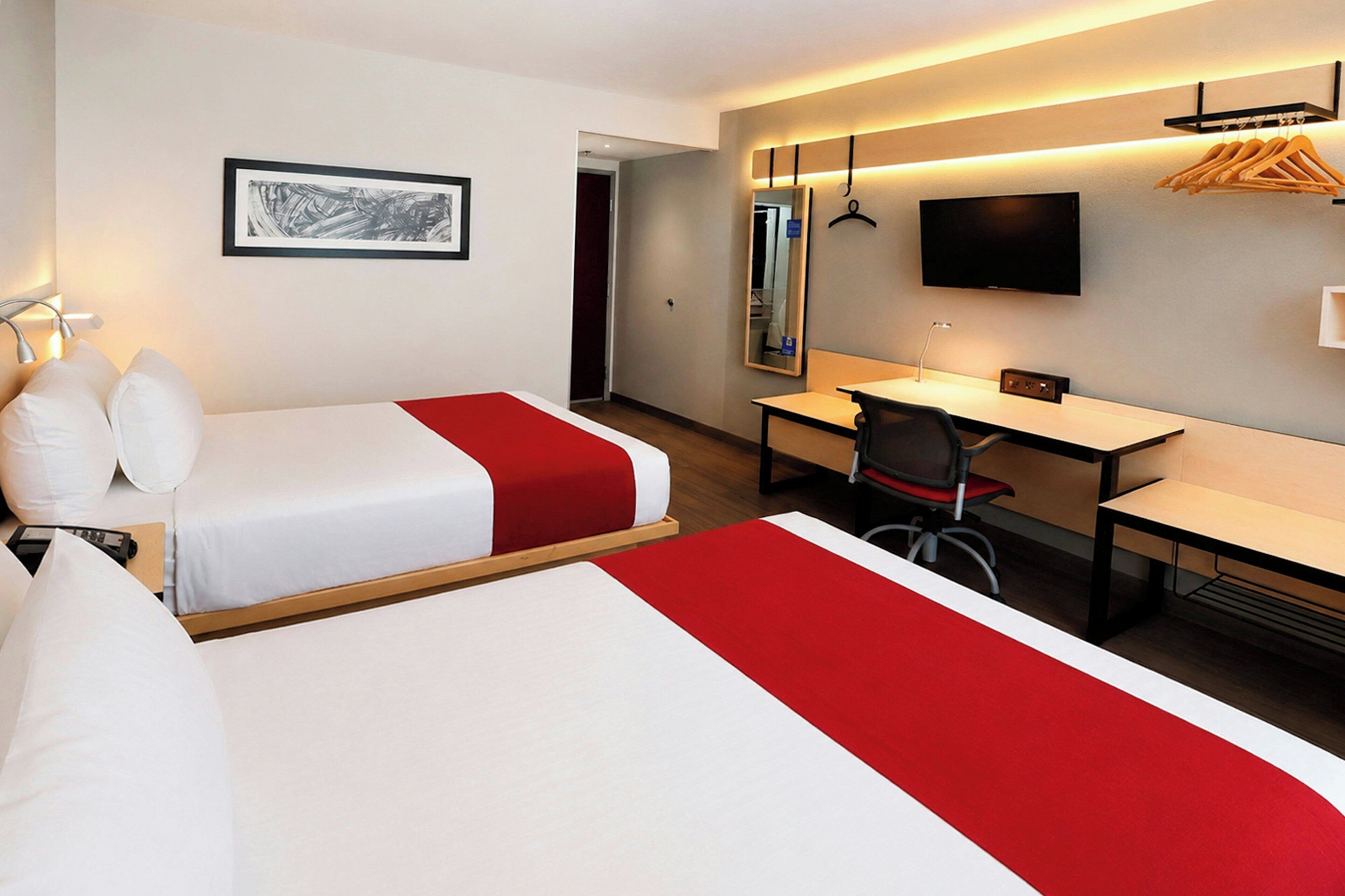 Quarto de hotel com duas camas, uma escrivaninha, uma cadeira, uma televisão e um espelho.