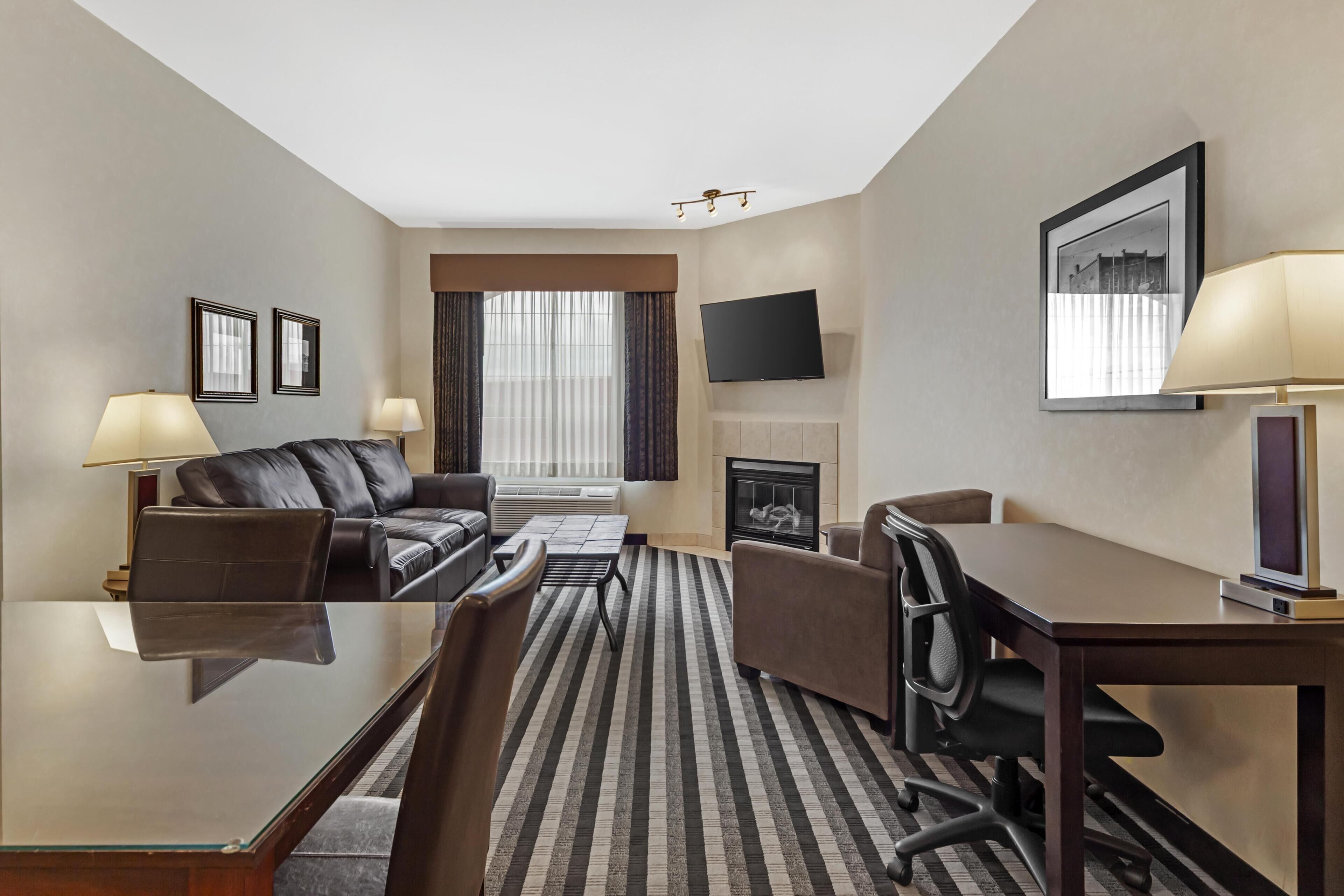 Quarto de hotel com sofá, escrivaninha, cadeira, televisão de tela plana, lareira e janela com cortinas.