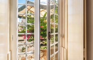 Visão através de uma porta de vidro com guarda-corrimão de metal, dando para uma área externa com plantas em vasos e uma cadeira branca.