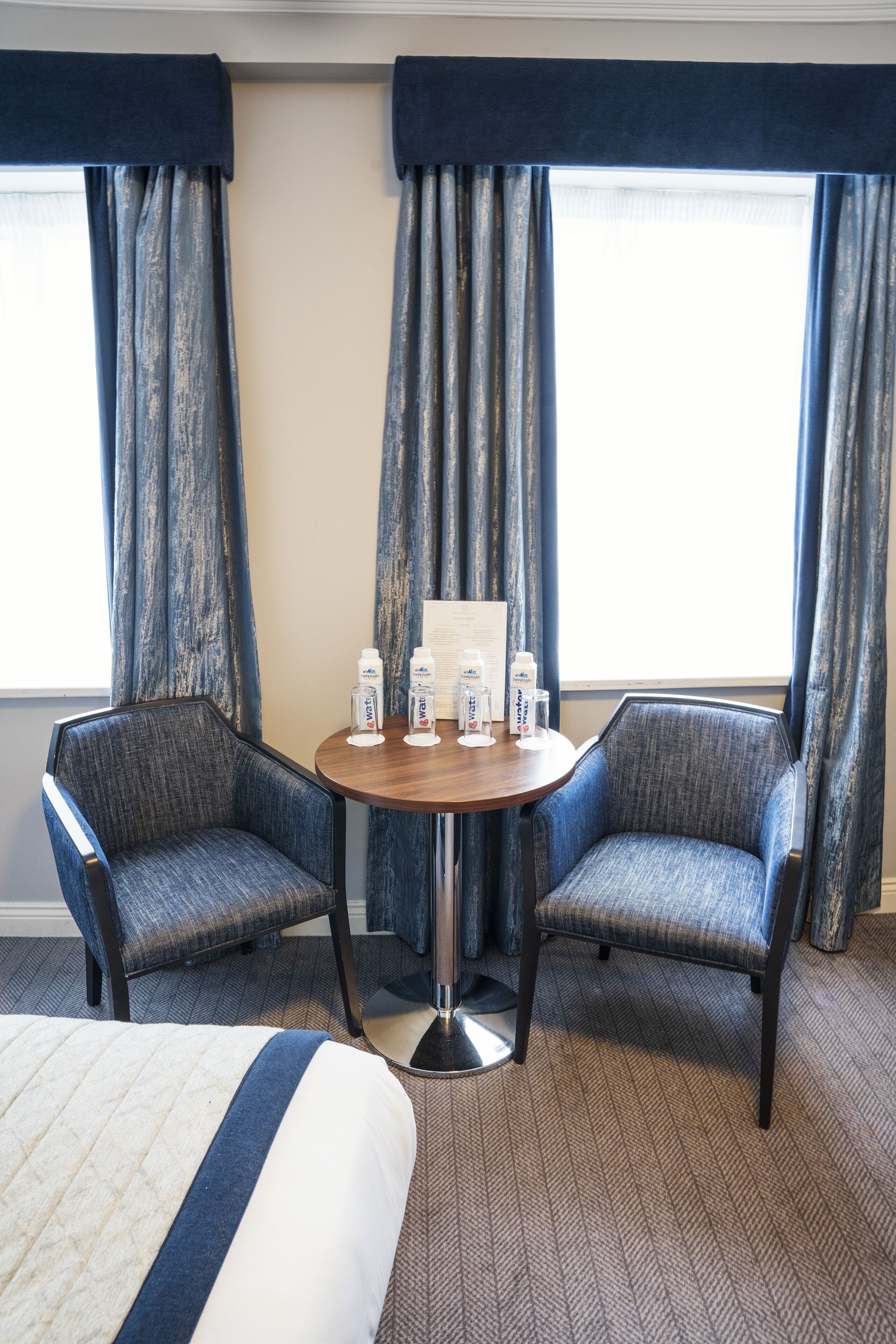 Quarto de hotel com cama, duas poltronas, mesa redonda pequena com artigos de higiene pessoal e janela com cortinas.