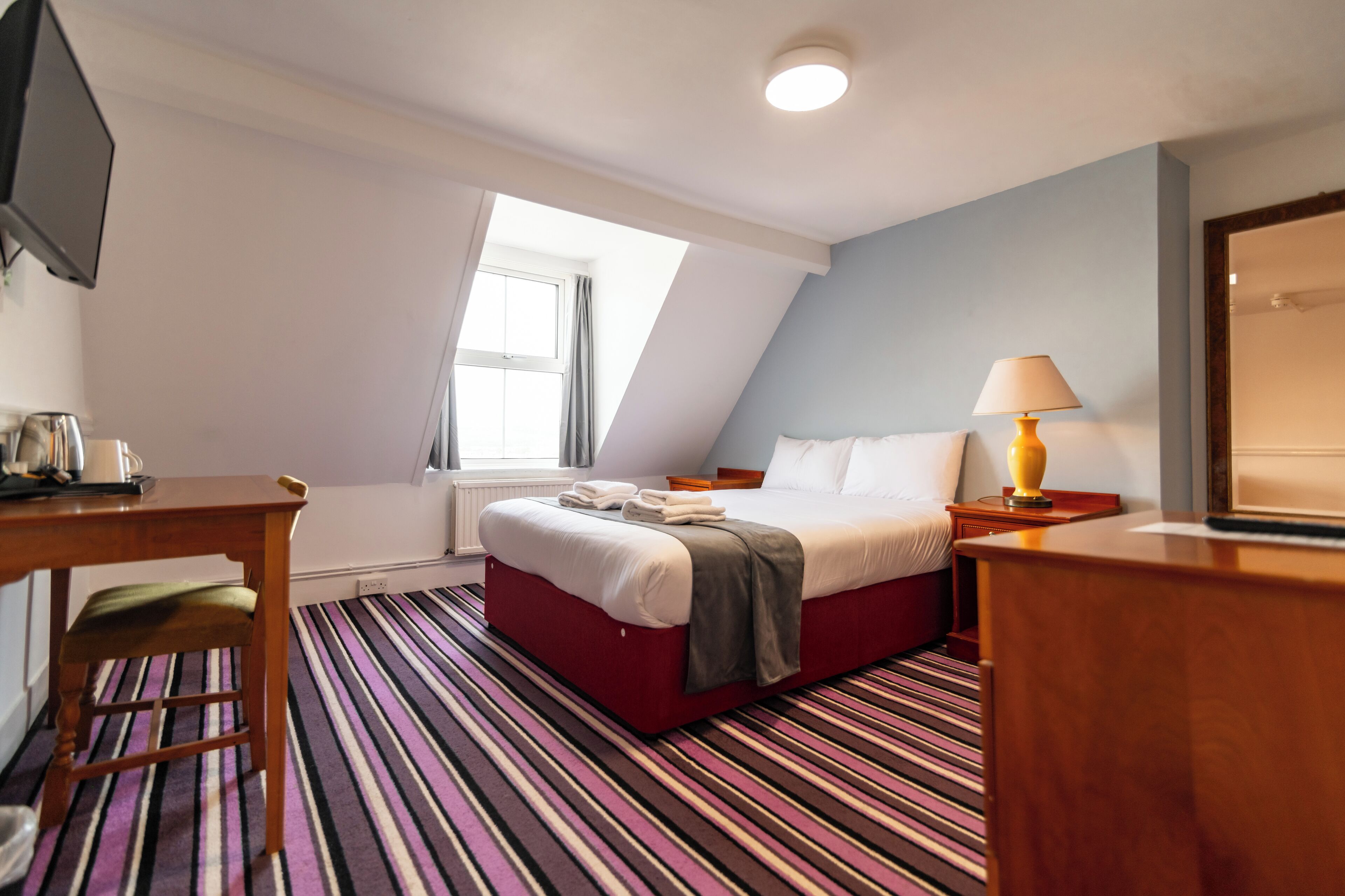 Quarto de hotel com cama, escrivaninha, cadeira, luminária e televisão.