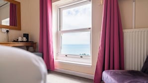 Quarto com vista para o mar, janela com cortinas rosa, escrivaninha de madeira com cafeteira e cadeira roxa.