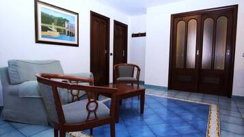 Uma sala de estar com um sofá, duas cadeiras de madeira, uma mesa, um quadro emoldurado e um conjunto de portas de madeira.
