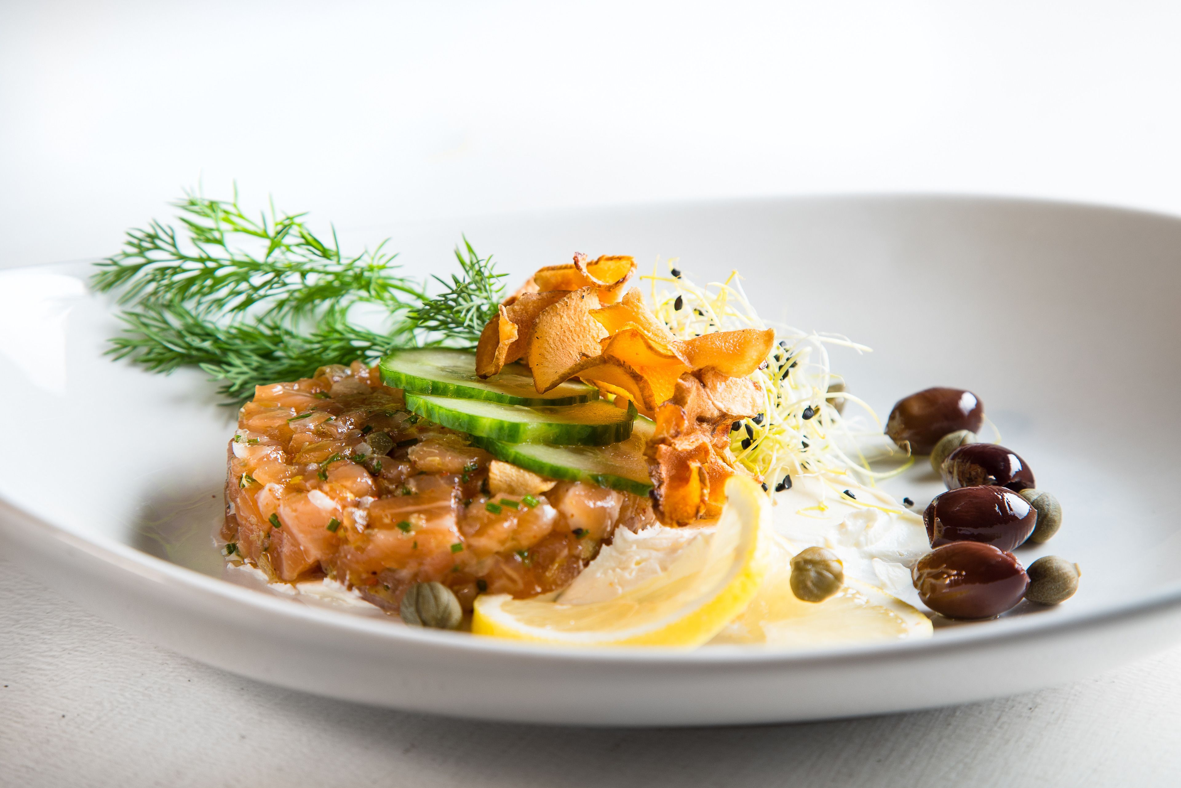 Um prato branco com uma porção de tartare de salmão, fatias de pepino, gomos de limão, alcaparras e um ramo de endro.