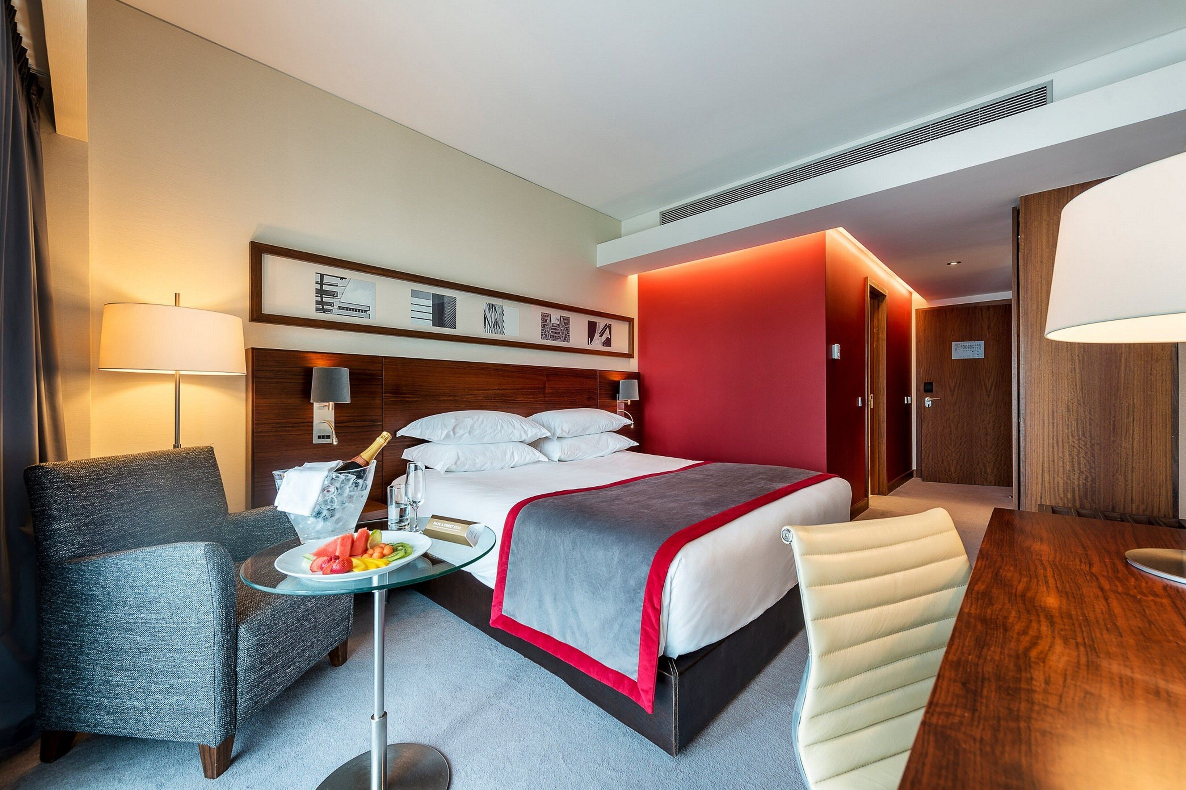 Quarto de hotel com cama, uma cadeira, mesa com um prato de fruta, luminária e escrivaninha de madeira.