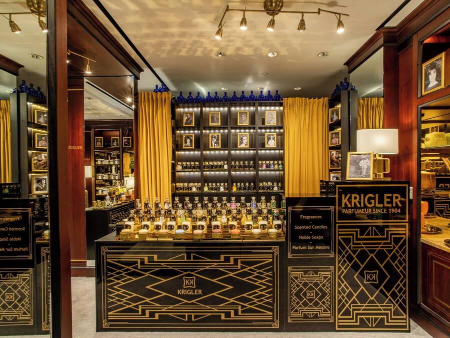 Interior de uma loja luxuosa com prateleiras de perfumes e velas, quadros emoldurados e um letreiro Krigler em destaque.
