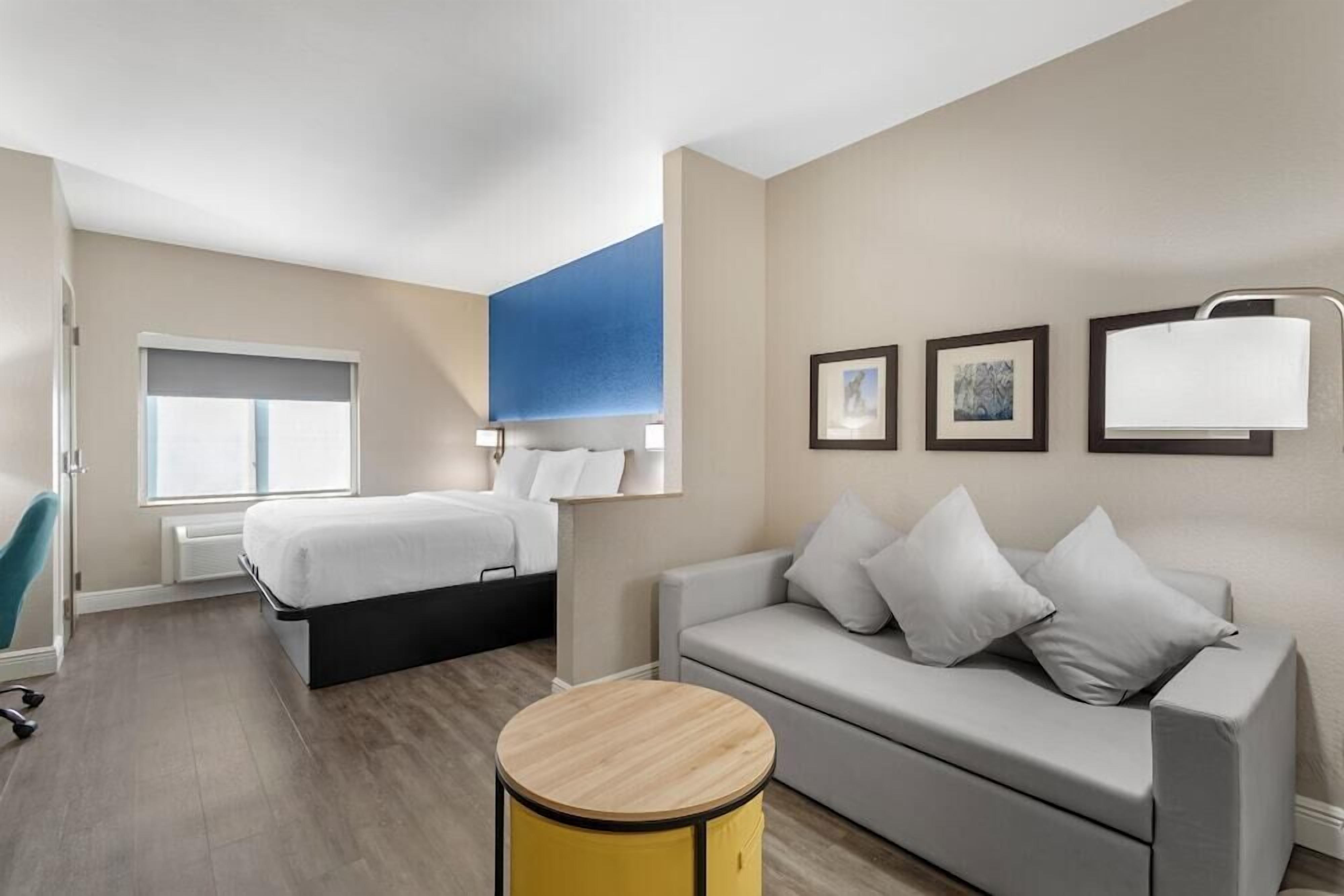 Quarto de hotel moderno com uma cama, um sofá, uma mesa de madeira redonda e uma janela com persianas.