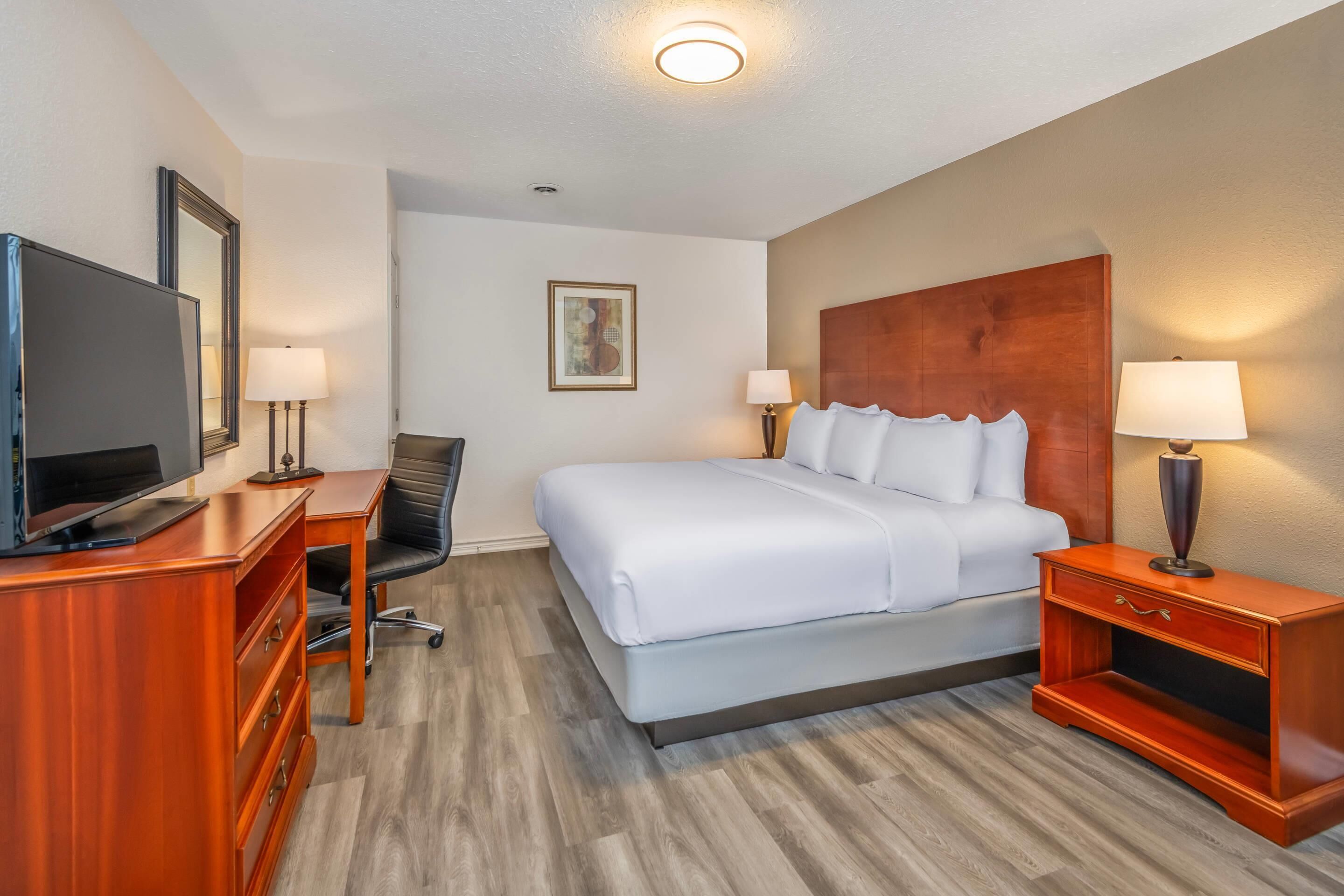 Quarto de hotel com cama, escrivaninha, cadeira, televisão e luminárias.