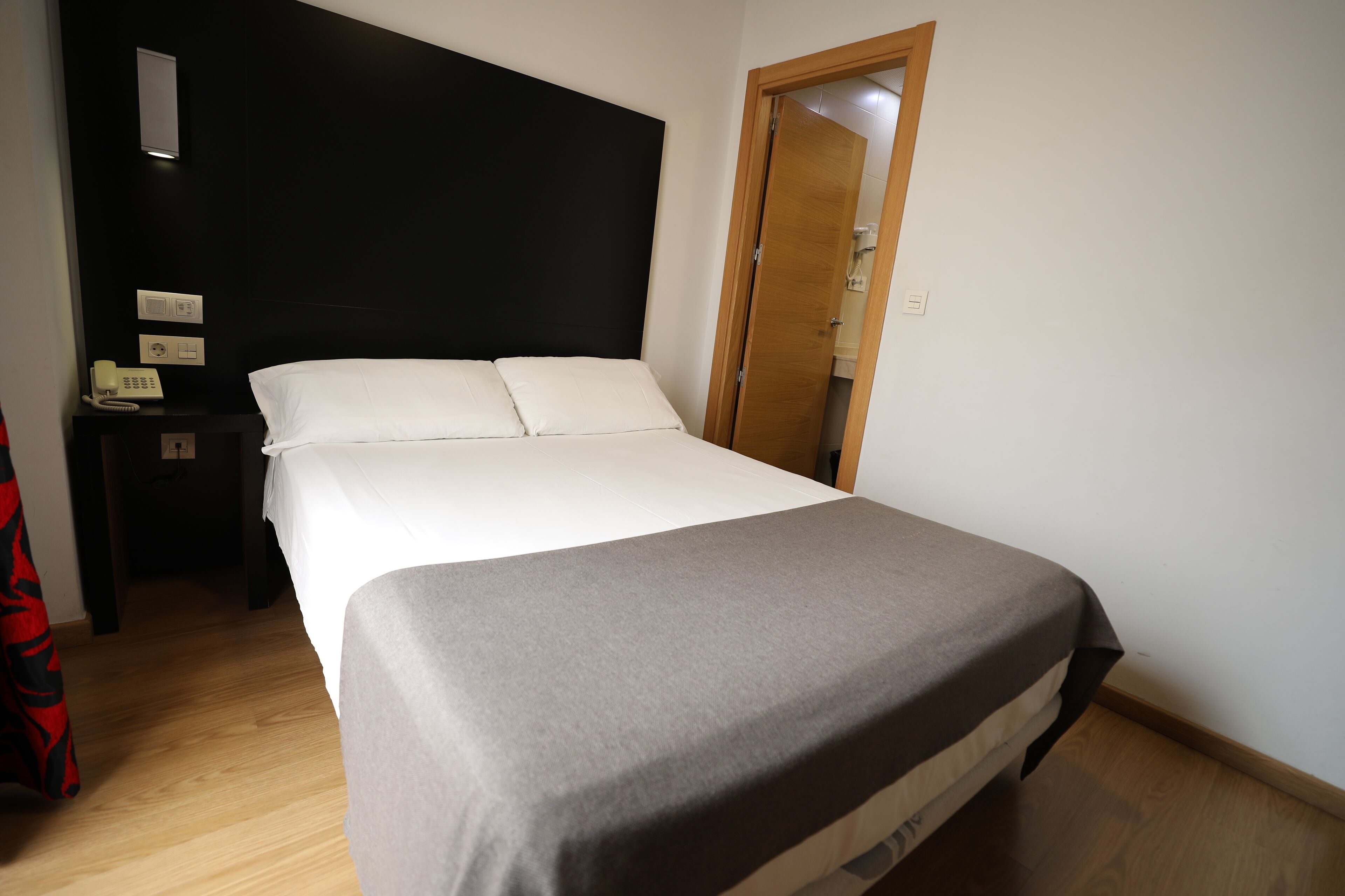 Cama bem arrumada com colcha cinza, mesa de cabeceira com telefone e cabeceira preta.
