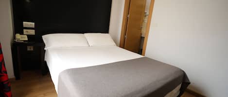 Cama bem arrumada com colcha cinza, mesa de cabeceira com telefone e cabeceira preta.