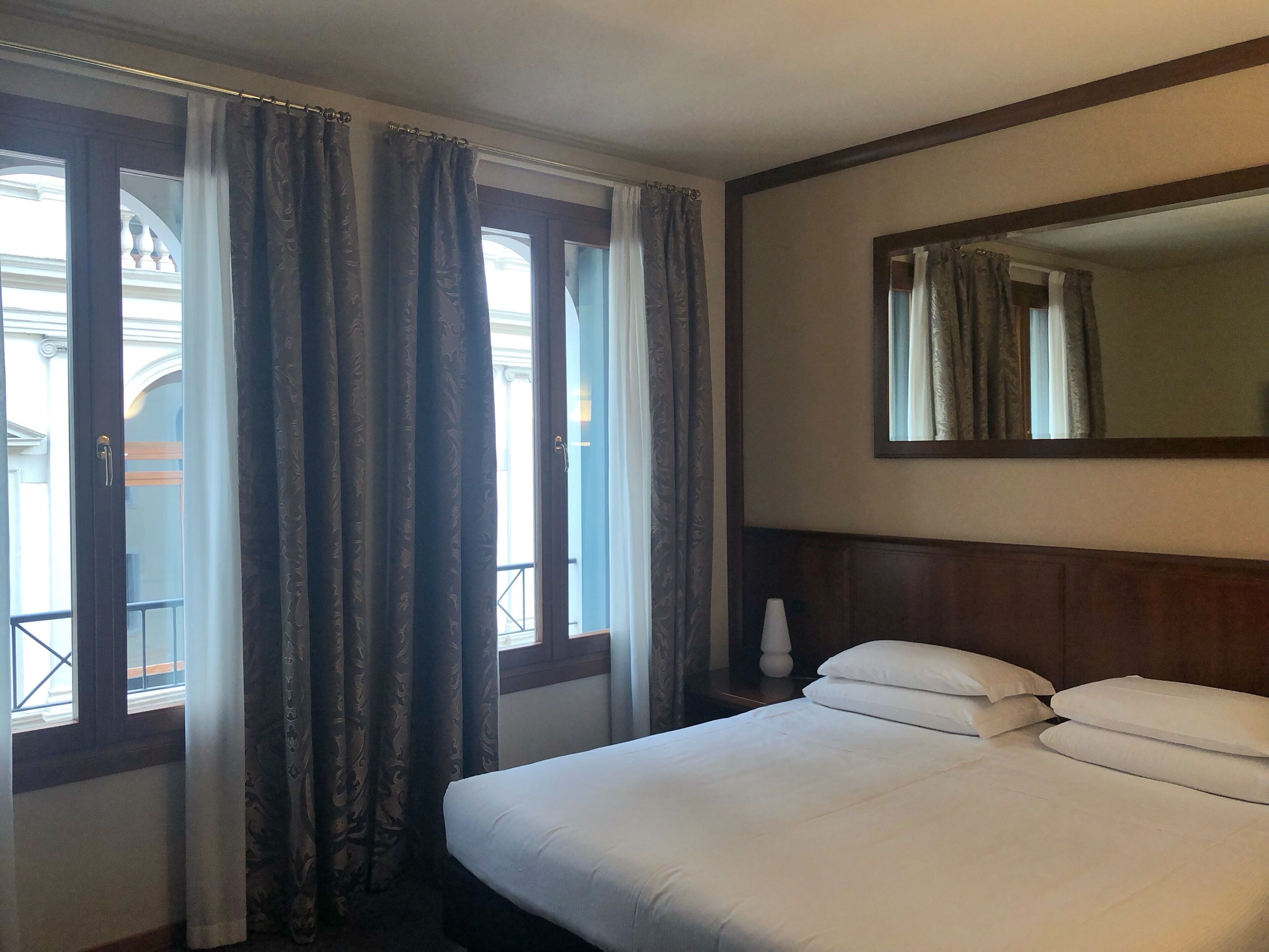 Quarto de hotel com uma cama, mesa de cabeceira, espelho e janela com cortinas.