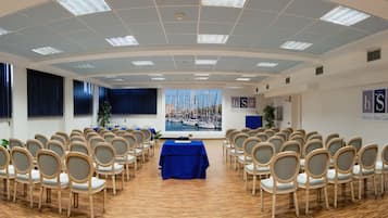 Sala de conferências com fileiras de cadeiras, uma mesa azul e uma janela grande com vista para um porto de iates.