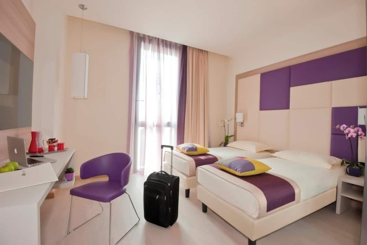 Quarto de hotel com cama, escrivaninha, cadeira, mala e planta em vaso.