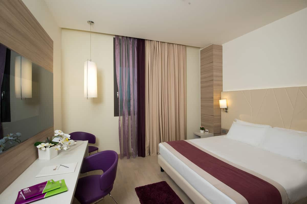 Quarto de hotel com cama, escrivaninha com cadeira, televisão e janela com cortinas.