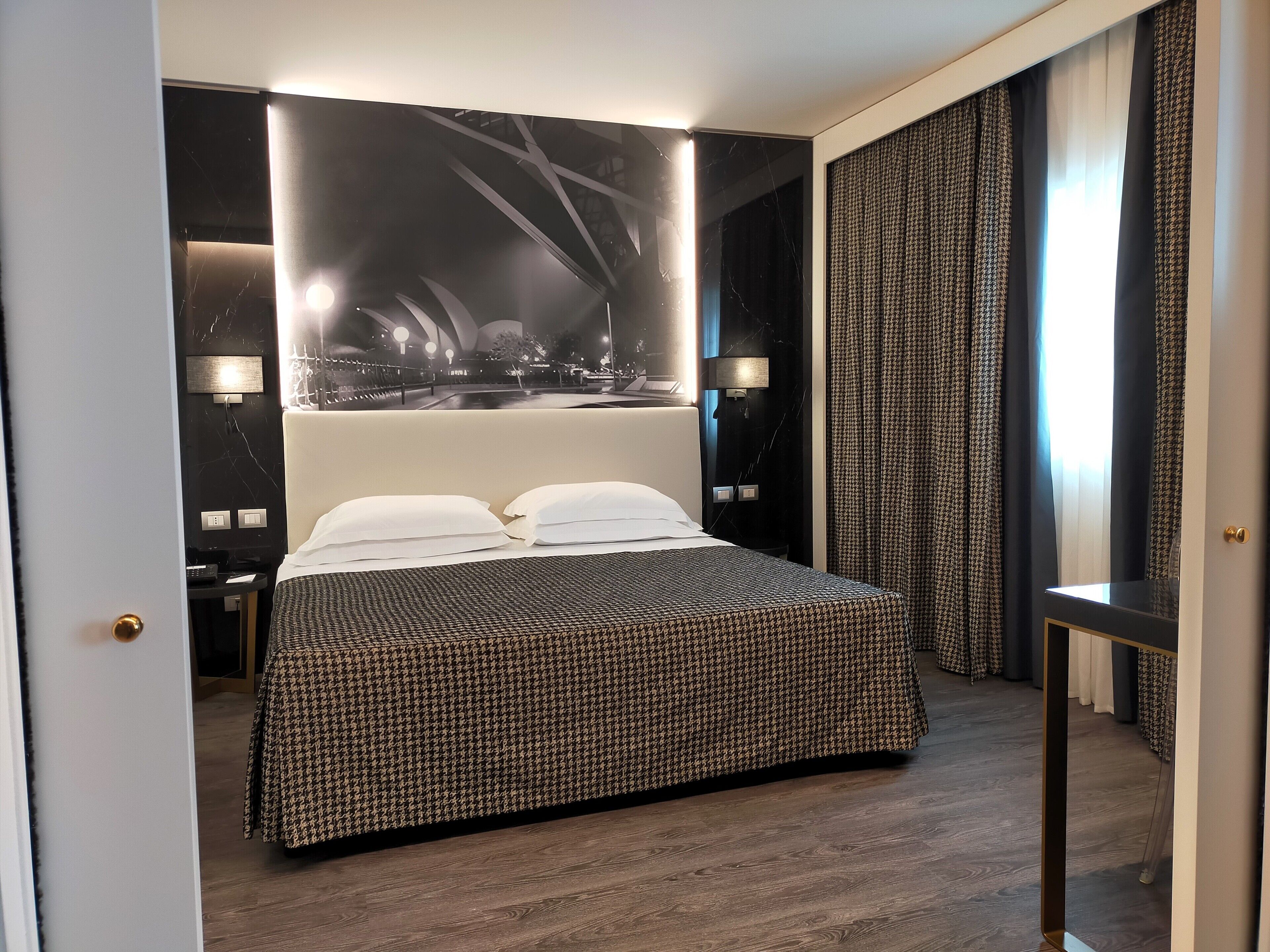 Quarto de hotel moderno com uma cama grande, um mural da paisagem urbana na parede e vista do céu noturno.