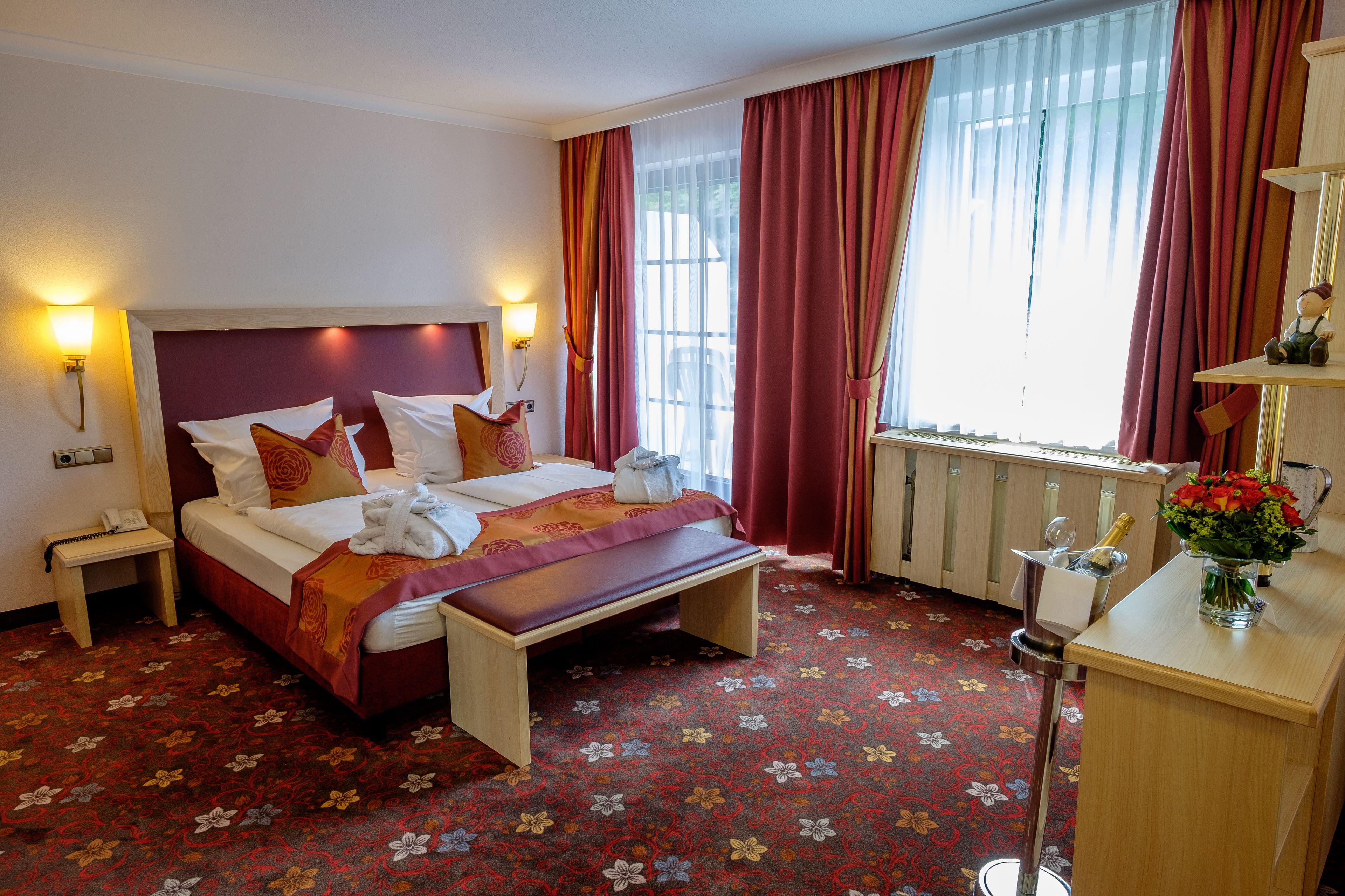 Quarto de hotel com cama, luminárias de cabeceira, mesa de cabeceira, escrivaninha com um vaso de flores e uma janela com cortinas.