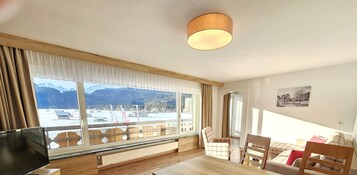 Quarto com uma grande janela oferecendo vista para as montanhas, piso de madeira, televisão, sofá e uma poltrona.
