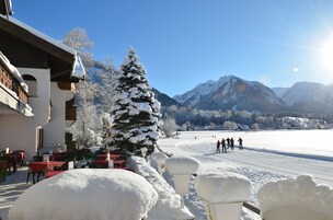Uma área externa com mesas e cadeiras coberta de neve, uma grande árvore coberta de neve e montanhas ao fundo.