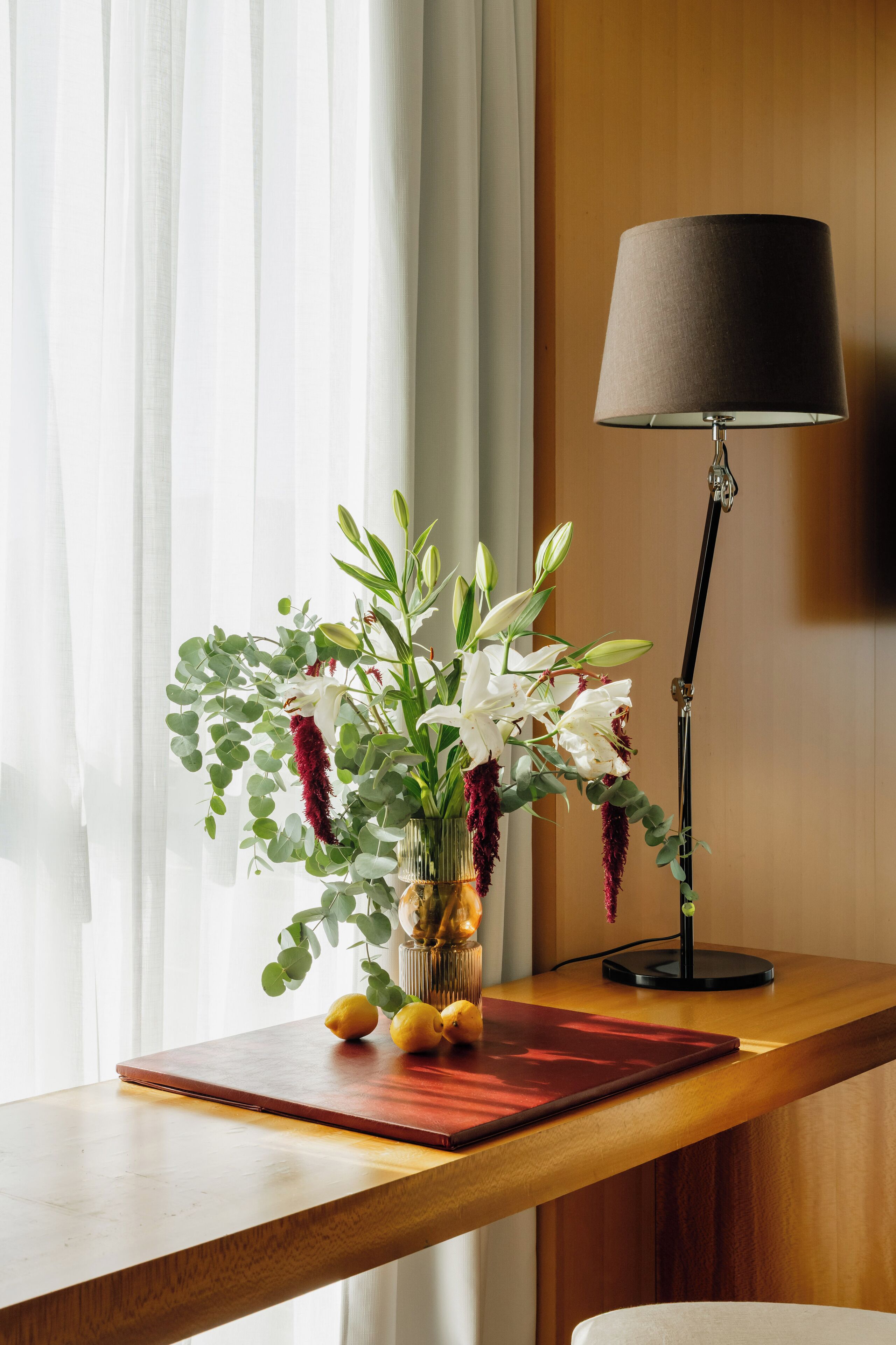 Uma mesa de madeira com um vaso de flores e limões, uma lâmpada e uma janela com cortinas leves.