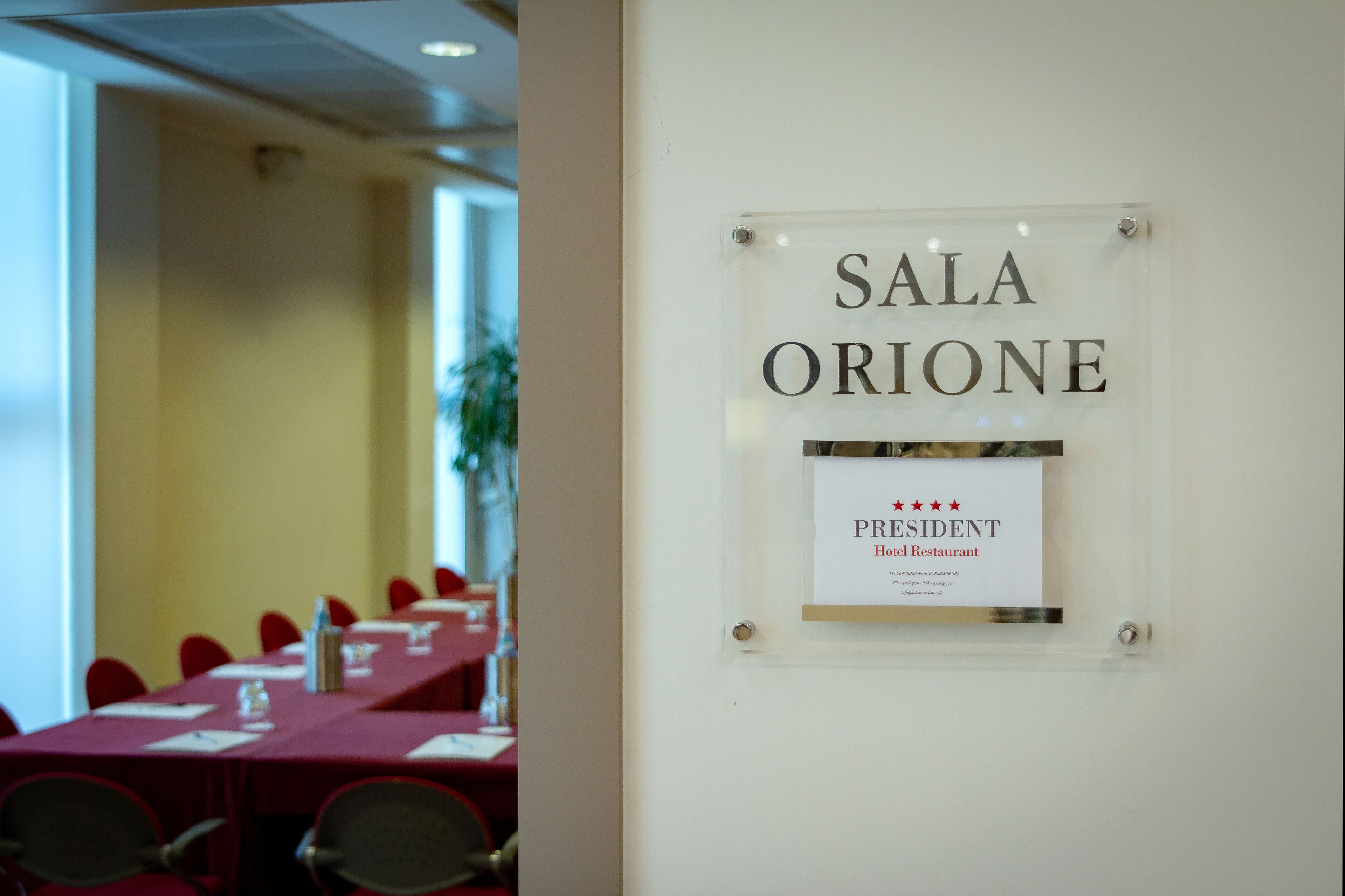 Uma sala de conferências com uma longa mesa e cadeiras, um letreiro emoldurado na parede com a inscrição "SALA ORIONE PRESIDENT Hotel Restaurant", e uma planta em vaso ao fundo.