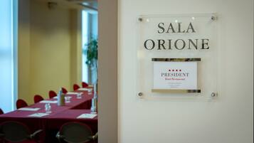 Uma sala de conferências com uma longa mesa e cadeiras, um letreiro emoldurado na parede com a inscrição "SALA ORIONE PRESIDENT Hotel Restaurant", e uma planta em vaso ao fundo.