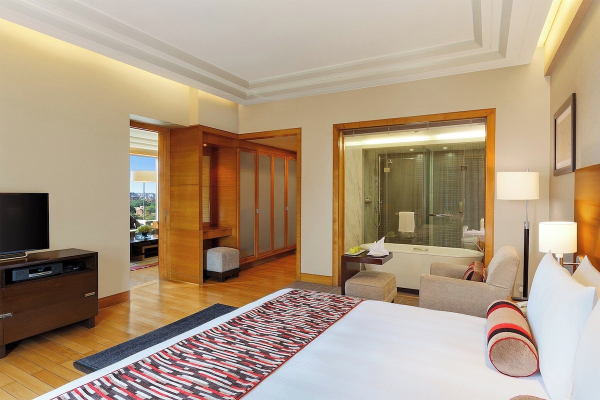 Quarto de hotel com cama, televisão, escrivaninha, cadeira e banheiro com espelho e chuveiro.