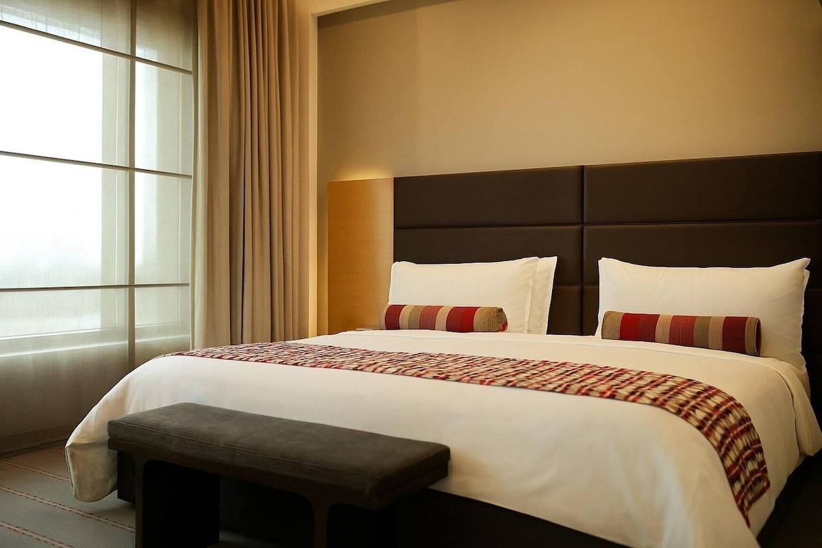 Quarto de hotel com uma cama grande, um banco, uma janela com cortinas e uma cabeceira.