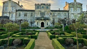 Um edifício histórico com fachada simétrica, entrada central e um jardim formal com sebes geometricamente aparadas.