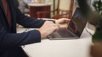 Uma pessoa usando um laptop em uma mesa, em um quarto com uma cadeira e uma mesinha ao fundo.