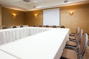 Sala de conferências com uma mesa comprida, cadeiras, tela de projetor e luminárias fixadas na parede.