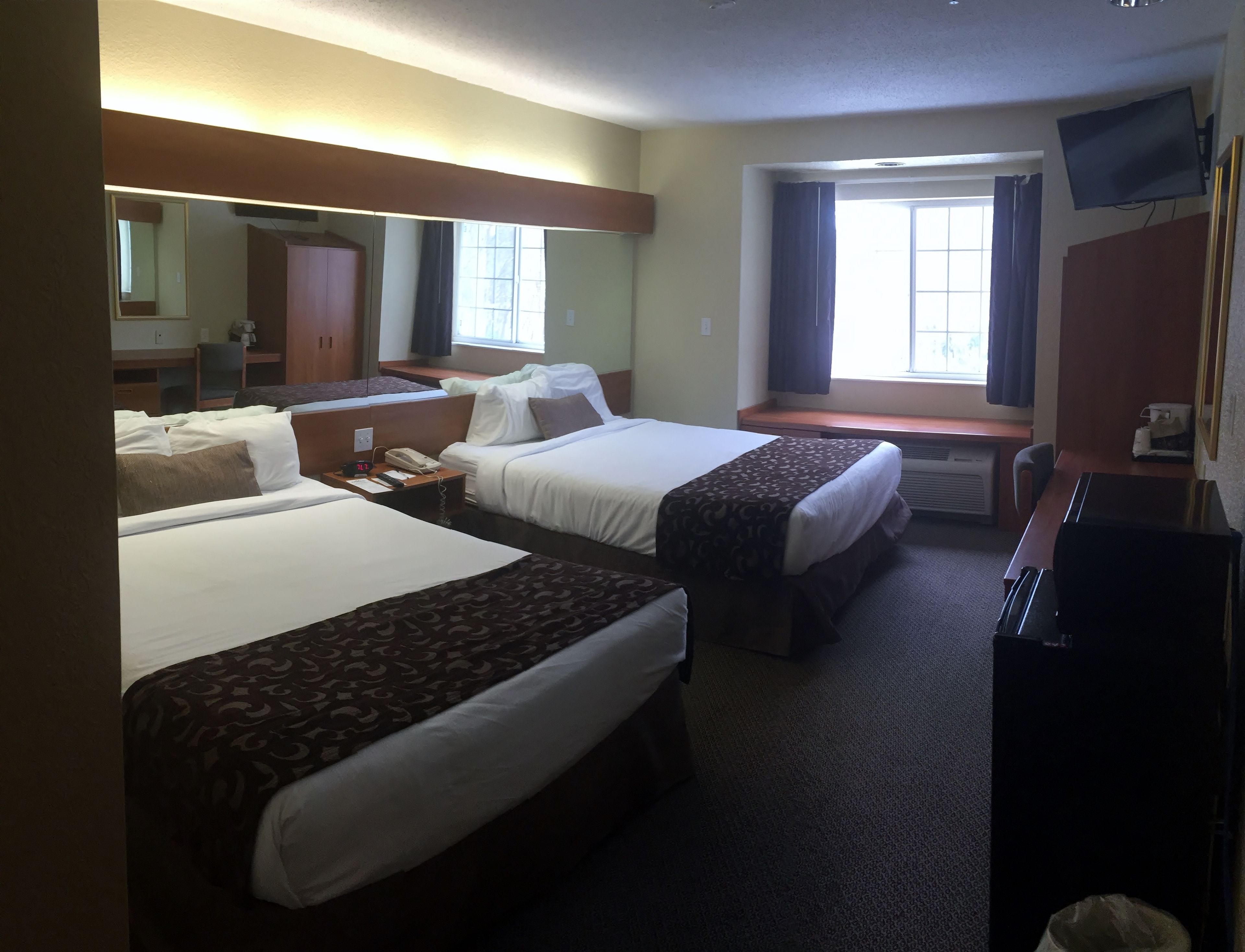 Quarto de hotel com duas camas, uma escrivaninha, uma cadeira, uma televisão e uma janela com cortinas.