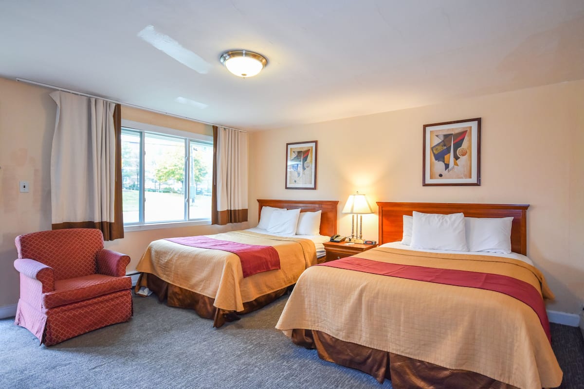 Quarto de hotel com duas camas, uma cadeira, um abajur e obras de arte nas paredes.