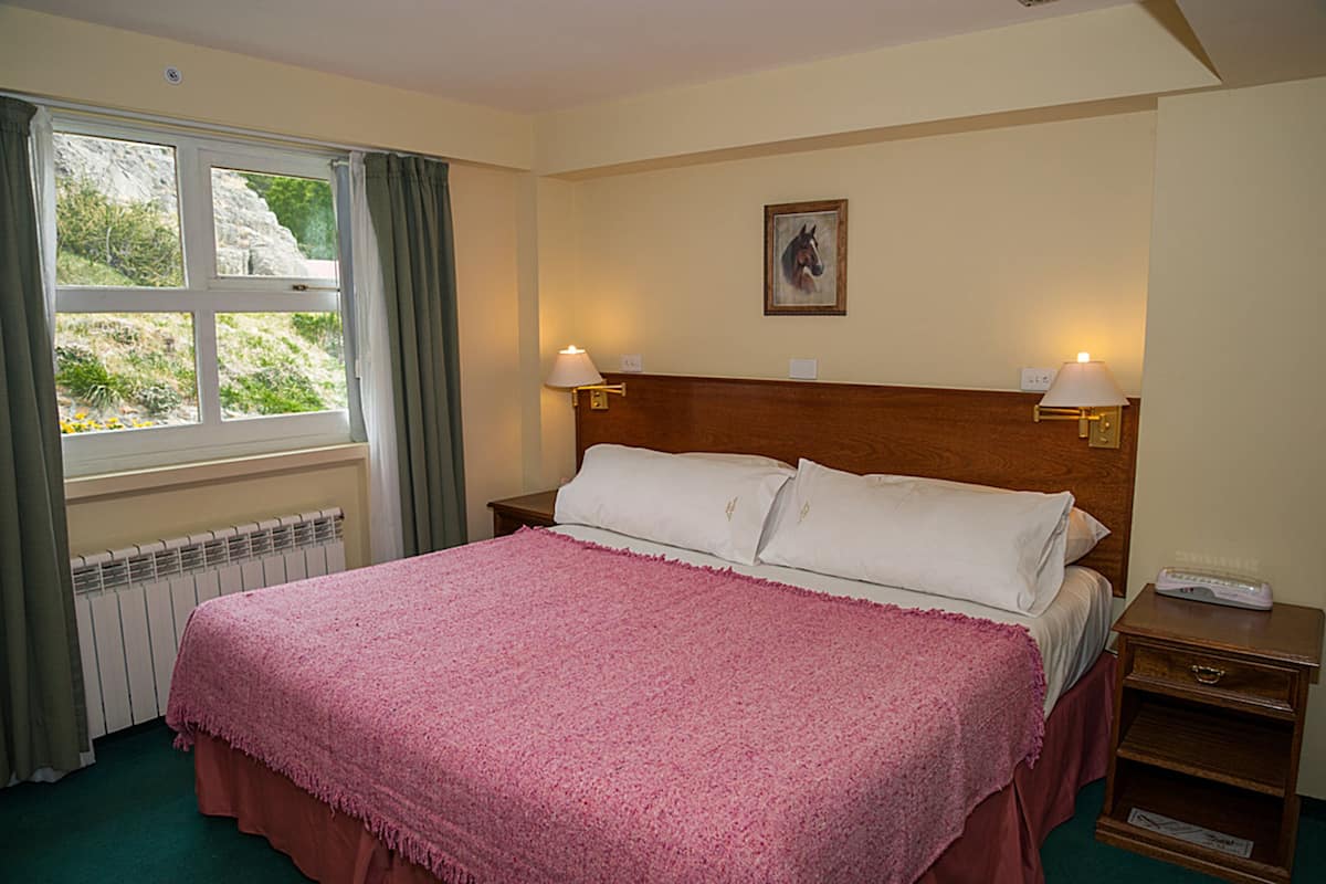 Quarto de hotel com cama, duas luminárias de cabeceira, mesa de apoio, quadro na parede e janela com cortinas.