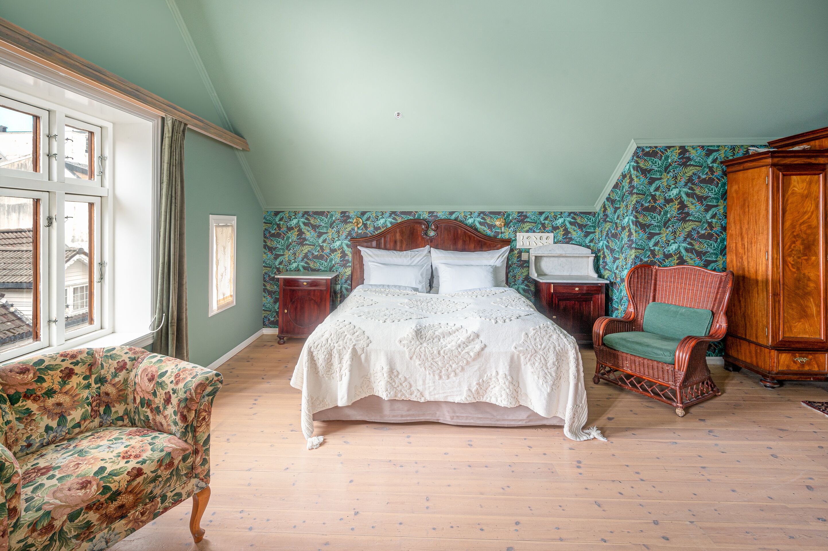 Quarto com uma cama grande, uma poltrona floral, uma cadeira de vime e um armário de madeira.