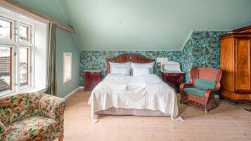 Quarto com uma cama grande, uma poltrona floral, uma cadeira de vime e um armário de madeira.