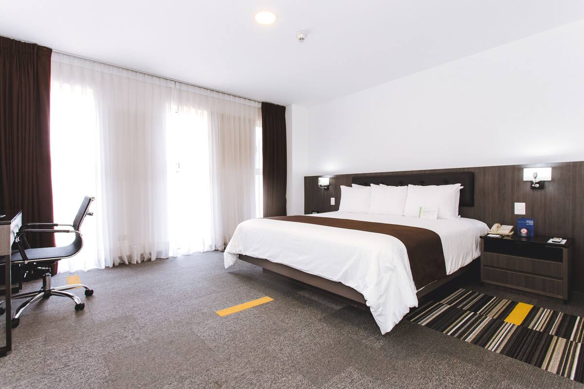 Quarto de hotel com uma cama grande, uma escrivaninha com cadeira, uma mesa de cabeceira com telefone e uma janela com cortinas.